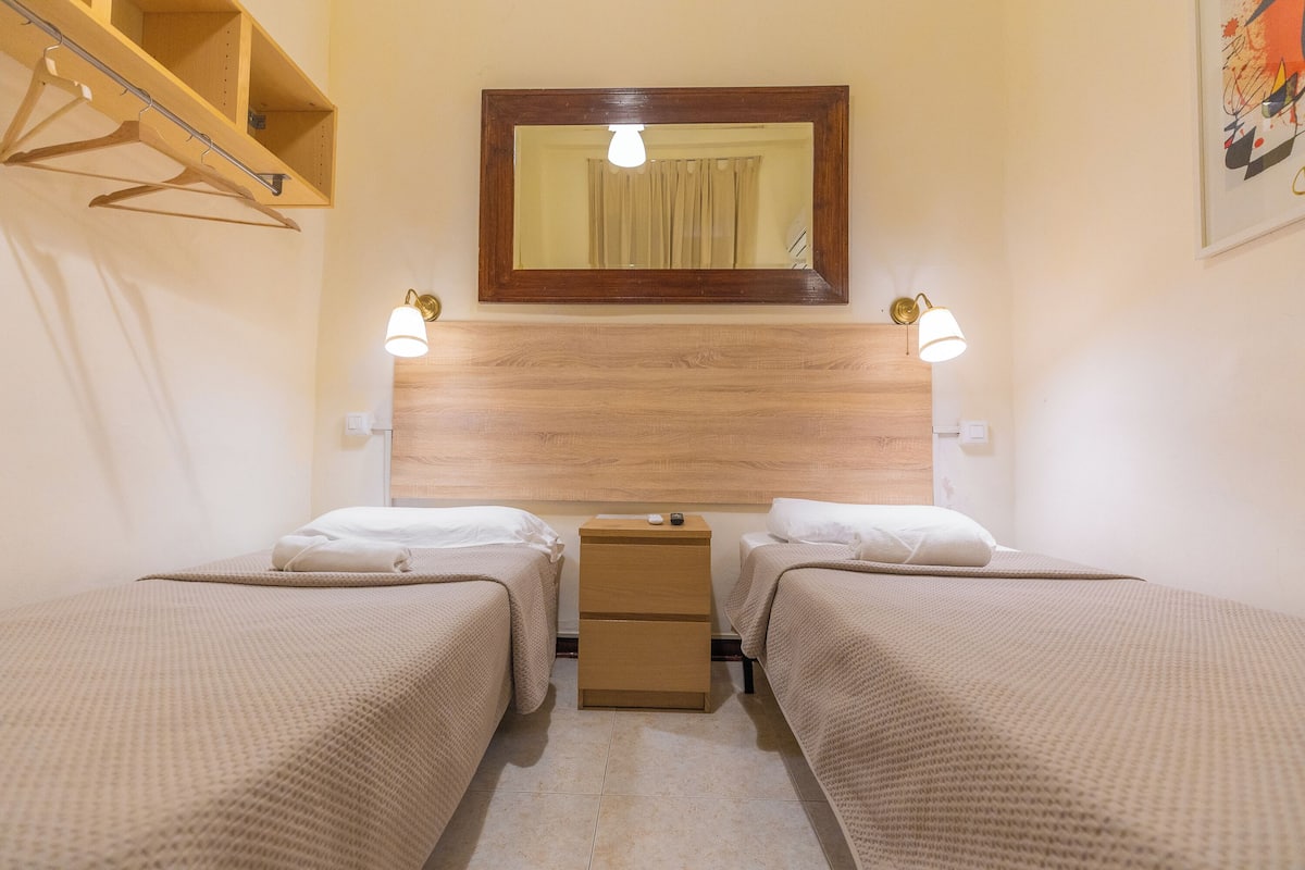 Quarto de hotel com duas camas de solteiro, cabeceira de madeira, mesinha de cabeceira com abajur e espelho sobre a cama.