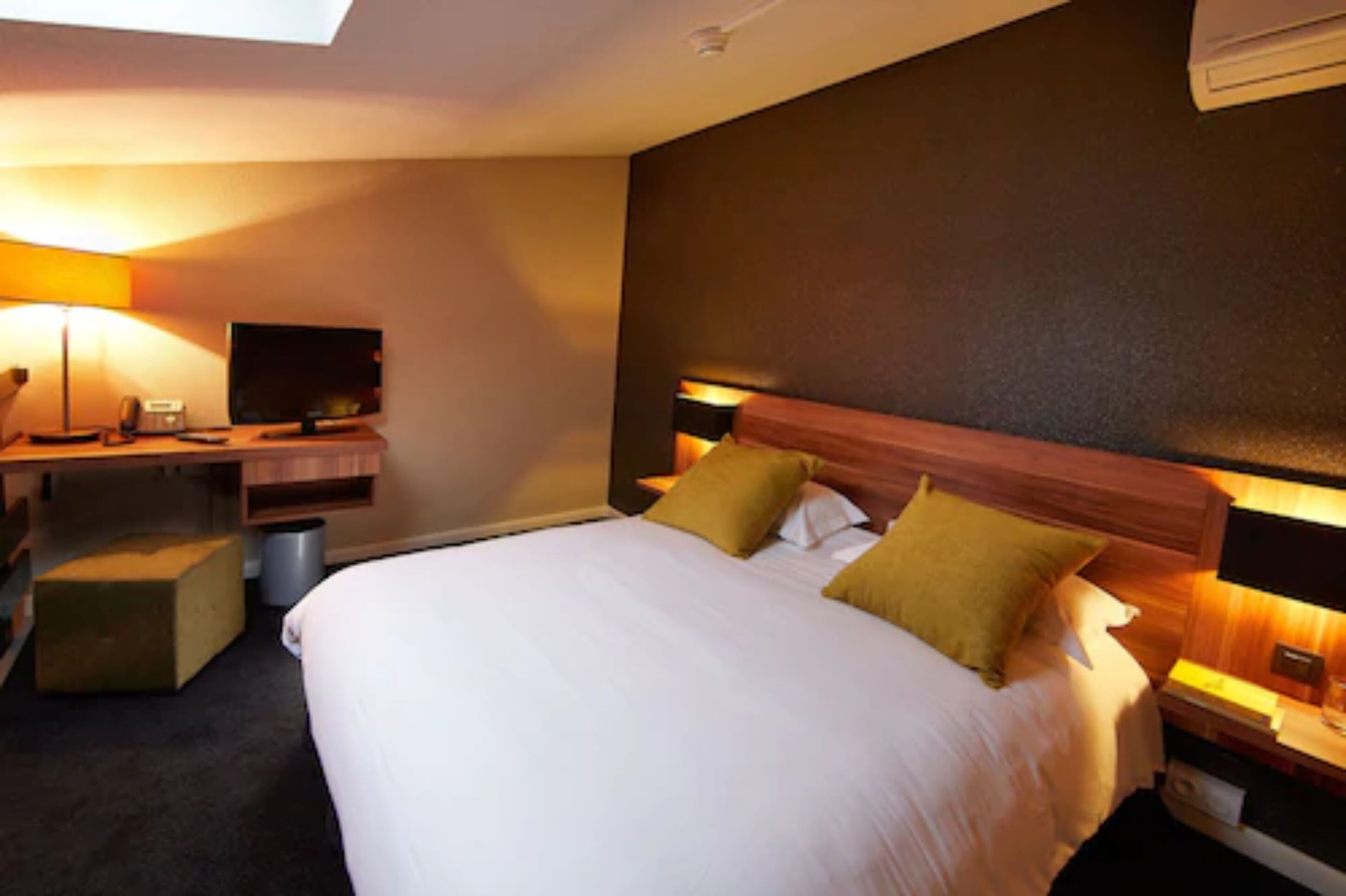 Quarto de hotel com cama, escrivaninha, computador, luminária e televisão.
