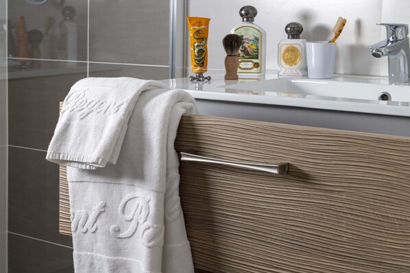 Um banheiro com uma toalha branca pendurada em um porta-toalhas de madeira, uma pia branca com um espelho acima e produtos de higiene no balcão da pia.