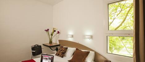 Quarto de hotel com uma cama, um laptop, um vaso com flores, uma mesa de cabeceira e uma janela com cortinas.