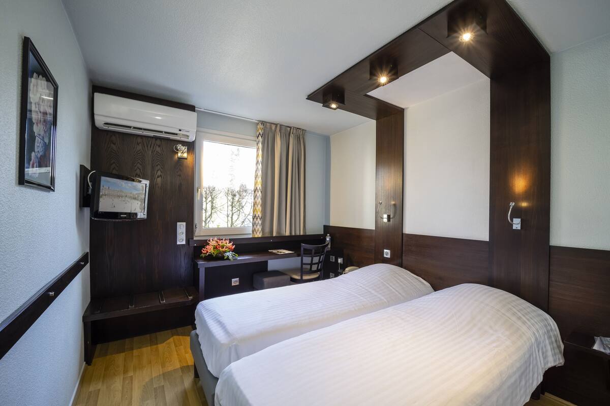 Quarto de hotel com duas camas, uma escrivaninha, uma cadeira, um espelho e uma janela com cortinas.
