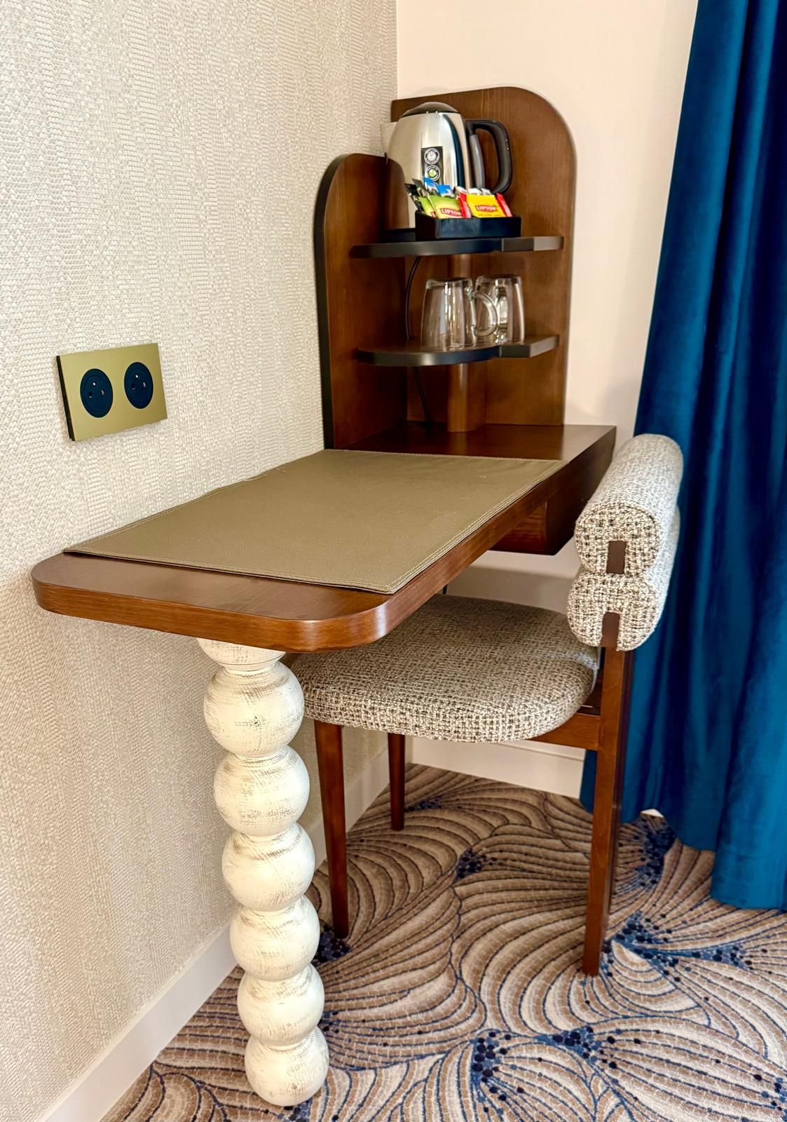 Canto de um quarto com uma mesa de madeira, uma cadeira com assento trançado, uma tomada e uma cafeteira em uma prateleira.