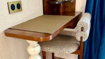 Canto de um quarto com uma mesa de madeira, uma cadeira com assento trançado, uma tomada e uma cafeteira em uma prateleira.