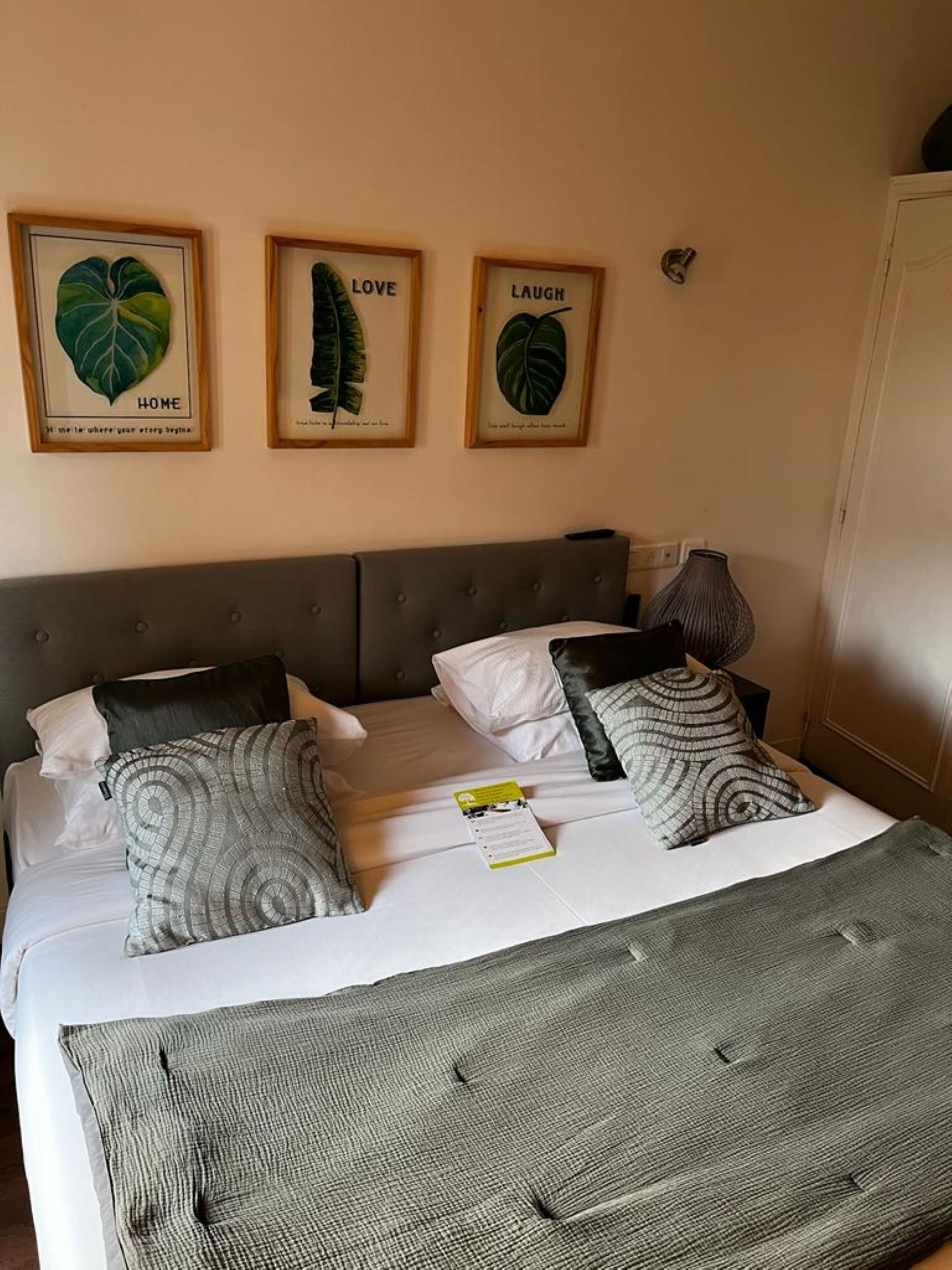 Uma cama bem arrumada com cabeceira cinza, várias almofadas e um edredom. Há três impressões emolduradas de folhas na parede acima da cama, cada uma com uma palavra diferente: HOME, LOVE, LAUGH.