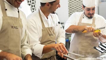 Cozinheiros de uniformes brancos e aventais preparam alimentos em uma cozinha com eletrodomésticos de aço inoxidável e parede estampada.