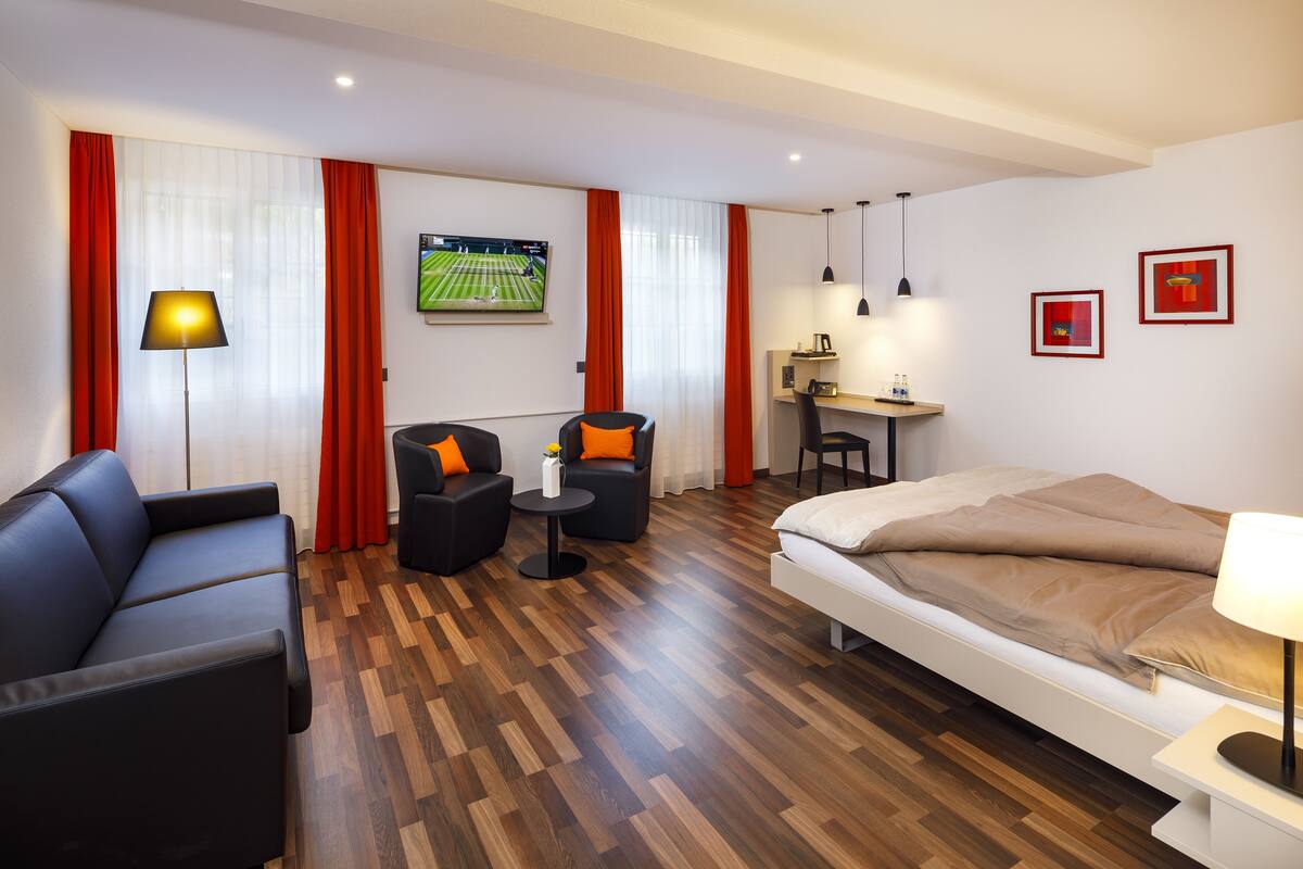 Quarto de hotel moderno com sofá, cama, escrivaninha e televisão exibindo um jogo esportivo.