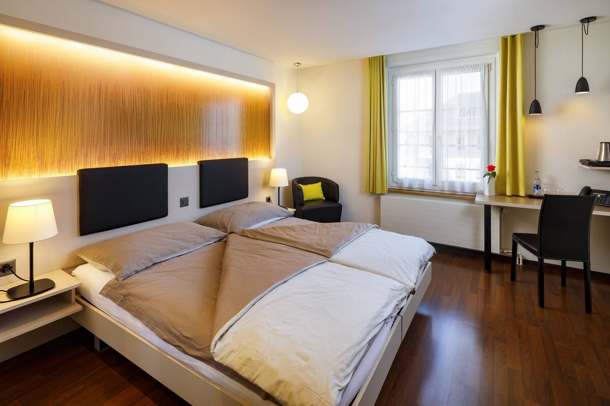 Quarto de hotel moderno com uma cama grande, mesas de cabeceira com luminárias, uma escrivaninha com cadeira e uma janela com cortinas.