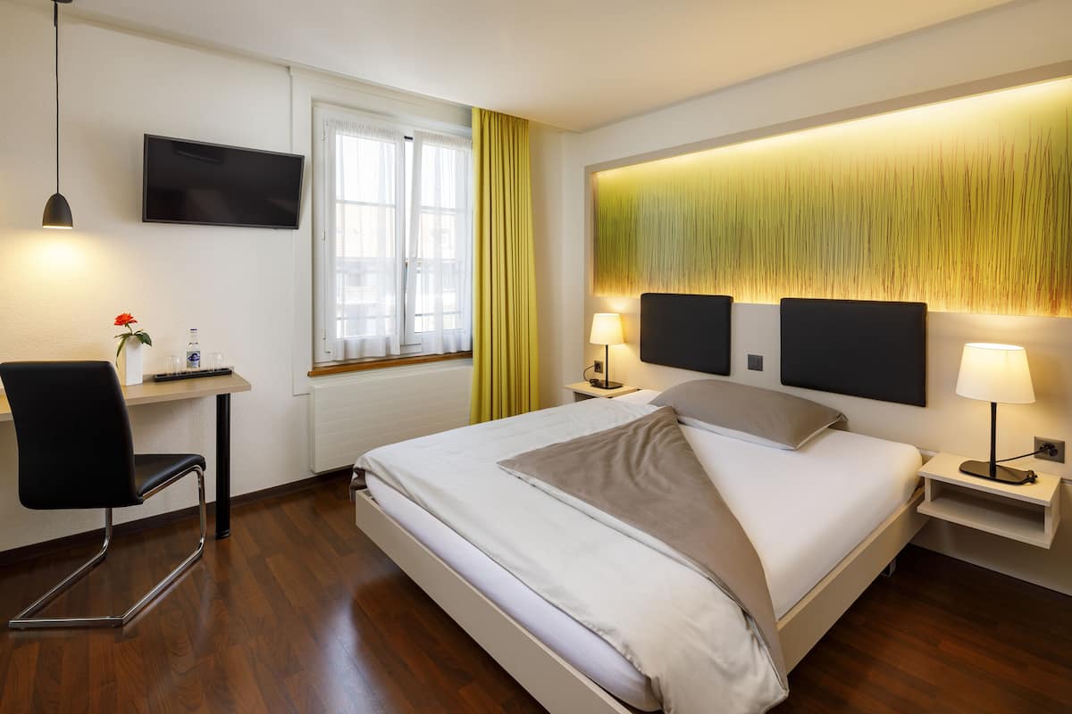 Quarto de hotel com cama, escrivaninha, cadeira, televisão e duas luminárias de cabeceira.
