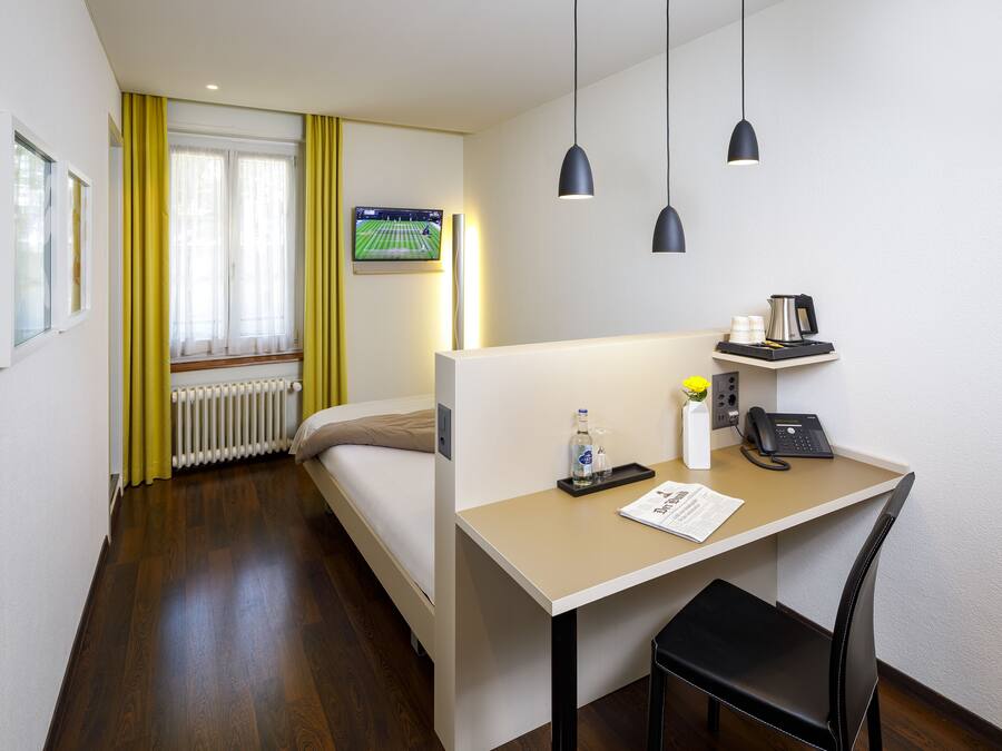 Quarto de hotel com cama, secretária com telefone e máquina de café, televisão na parede e janela com cortinas.