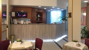 Uma área de recepção de hotel com balcão, televisão e área de estar com mesas e cadeiras.