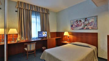 Quarto de hotel com cama, escrivaninha, cadeira, computador e uma obra de arte na parede.