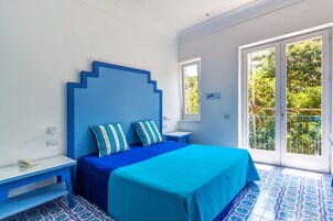 Quarto com cama azul, almofadas listradas, mesa de cabeceira branca, janela com vista para o verde e piso ladrilhado.