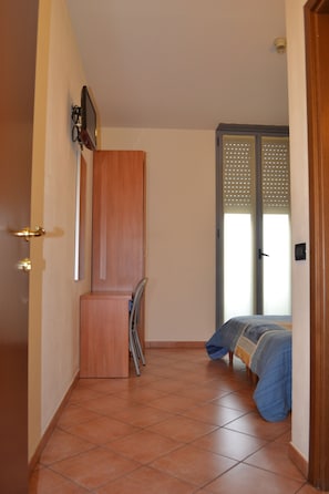 Um corredor estreito com um armário de madeira, uma cama com colcha azul e uma porta de vidro deslizante com padrão de treliça.