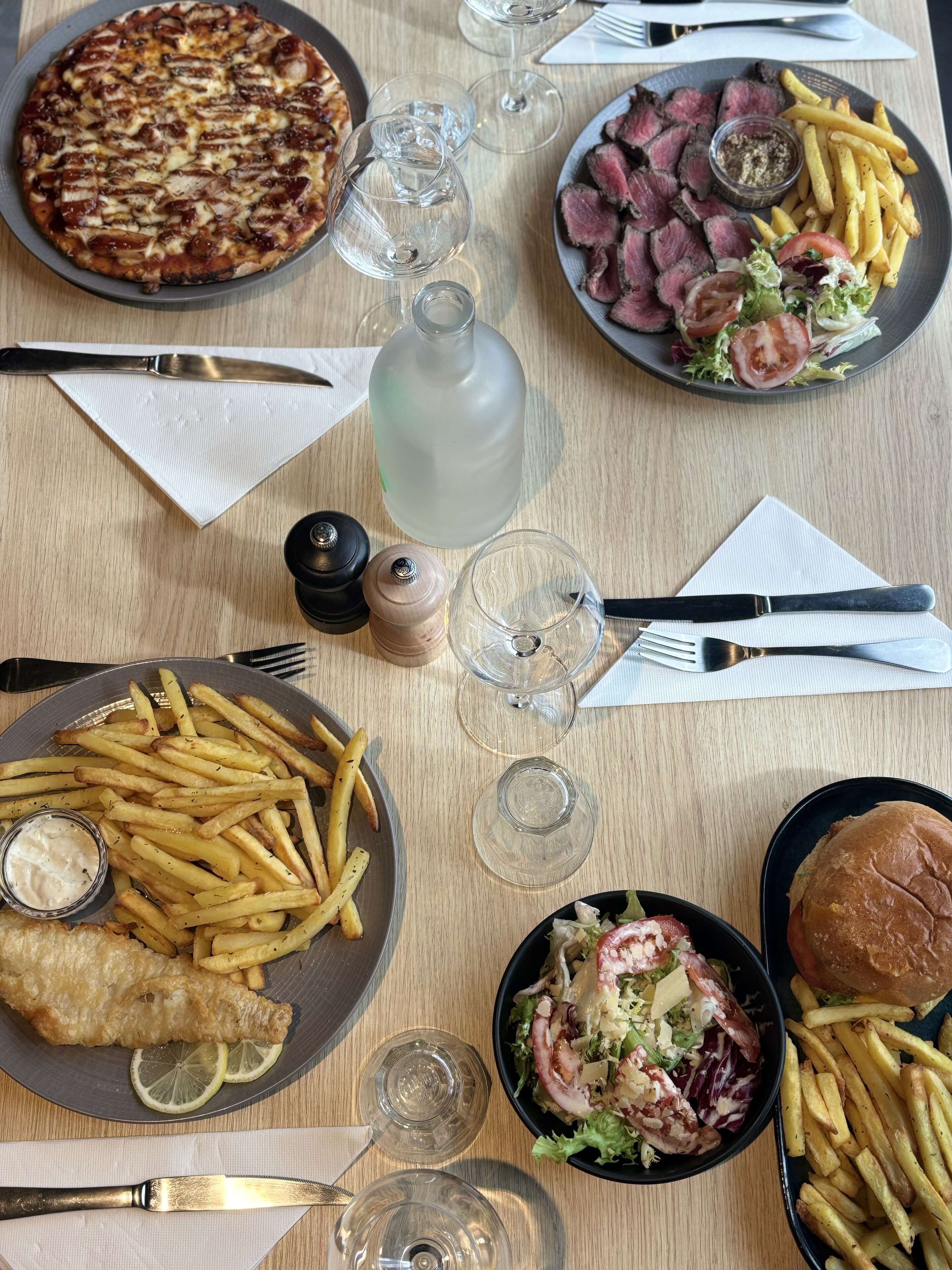 Uma mesa posta com diversos pratos, incluindo pizza, bife, batatas fritas, peixe com batatas e uma salada.