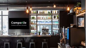 Uma área de bar moderna com uma tela grande exibindo "Campanile Créateurs de bons moments". Há banquetas, uma máquina de café e prateleiras com garrafas e copos.