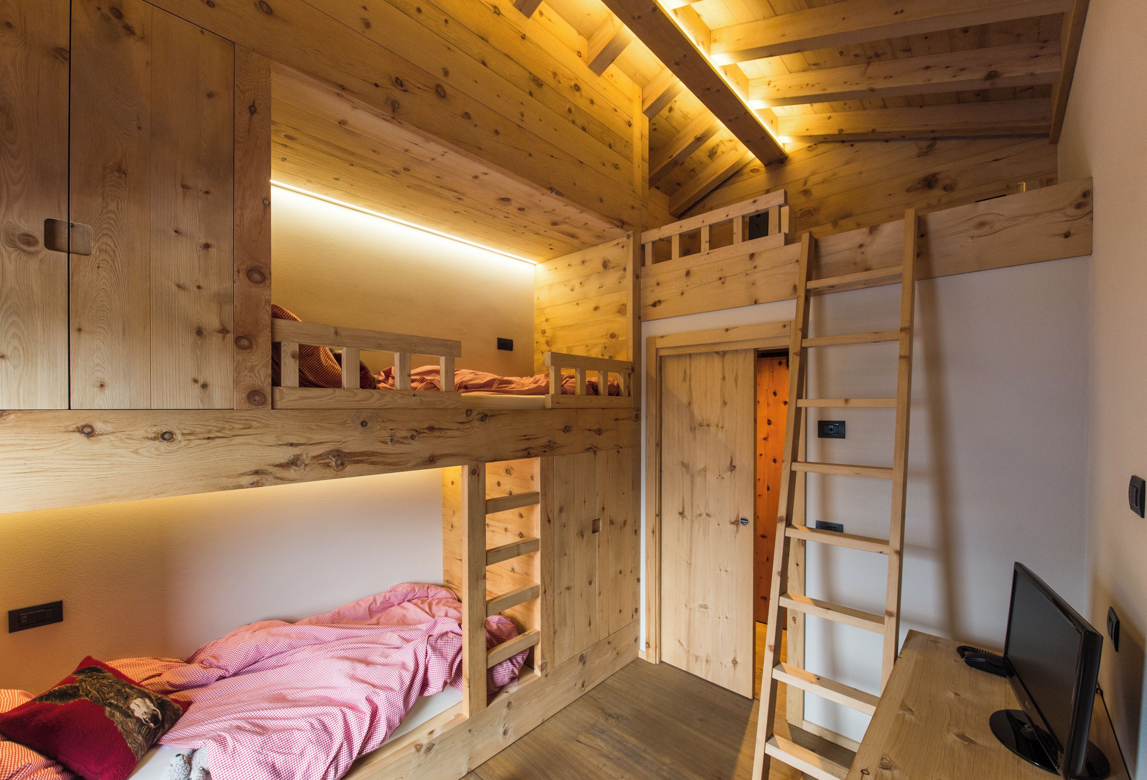 Interior de uma cabana de madeira com beliches, uma escada e uma escrivaninha com um computador.