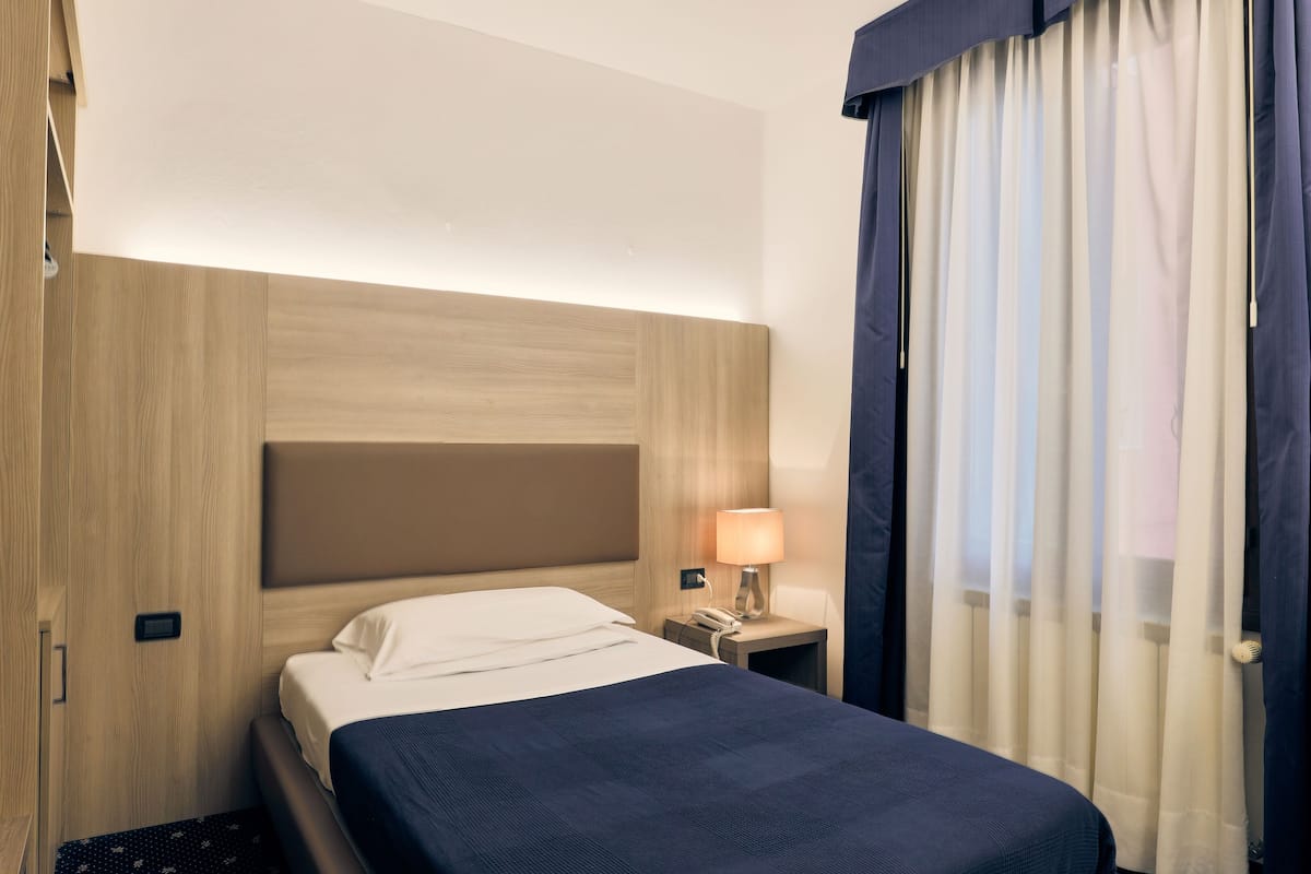 Quarto de hotel com cabeceira de madeira, cama com colcha azul escura, luminária de cabeceira e janela com cortinas finas.