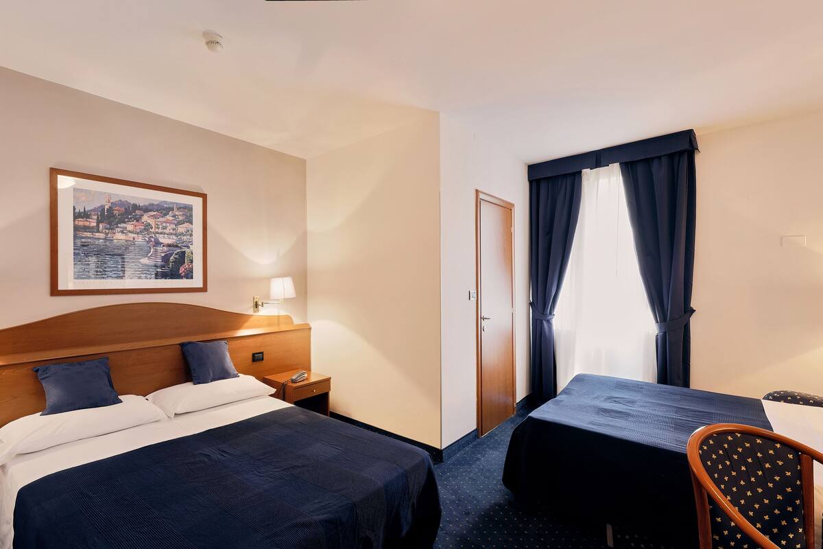 Quarto de hotel com cabeceira de madeira, duas camas, uma escrivaninha e uma cadeira. Há um quadro na parede, uma janela com cortinas e uma porta.
