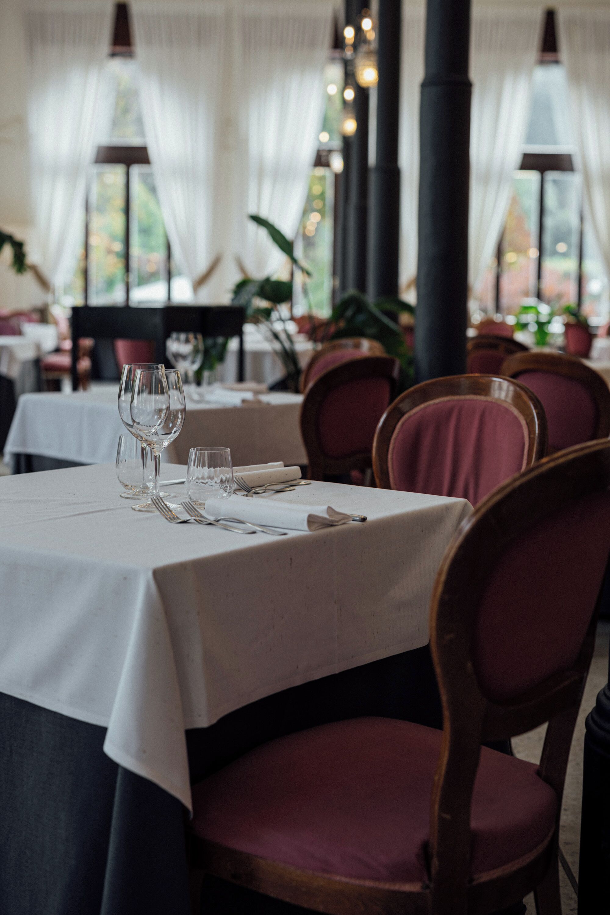 Interior de restaurante com mesas postas para refeições, cadeiras e vista através de grandes janelas.
