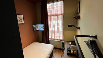 Quarto de hotel com cama, televisão na parede, janela com persianas, escrivaninha com luminária e uma cadeira.
