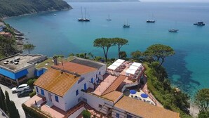 Um edifício costeiro com terraço virado para o mar, com vários barcos ao fundo e uma árvore em primeiro plano.