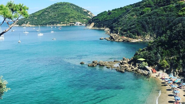 Uma área costeira com águas turquesa cristalinas, uma praia de areia e afloramentos rochosos.