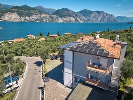 Um edifício moderno com painéis solares no telhado, entrada com calçada e vista para o mar e montanhas ao fundo.