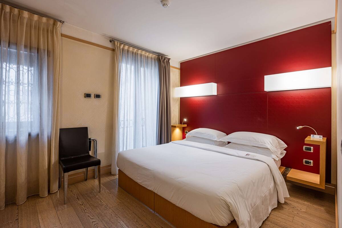 Quarto de hotel com cama grande, uma cadeira, mesa de cabeceira e parede com detalhes vermelhos.