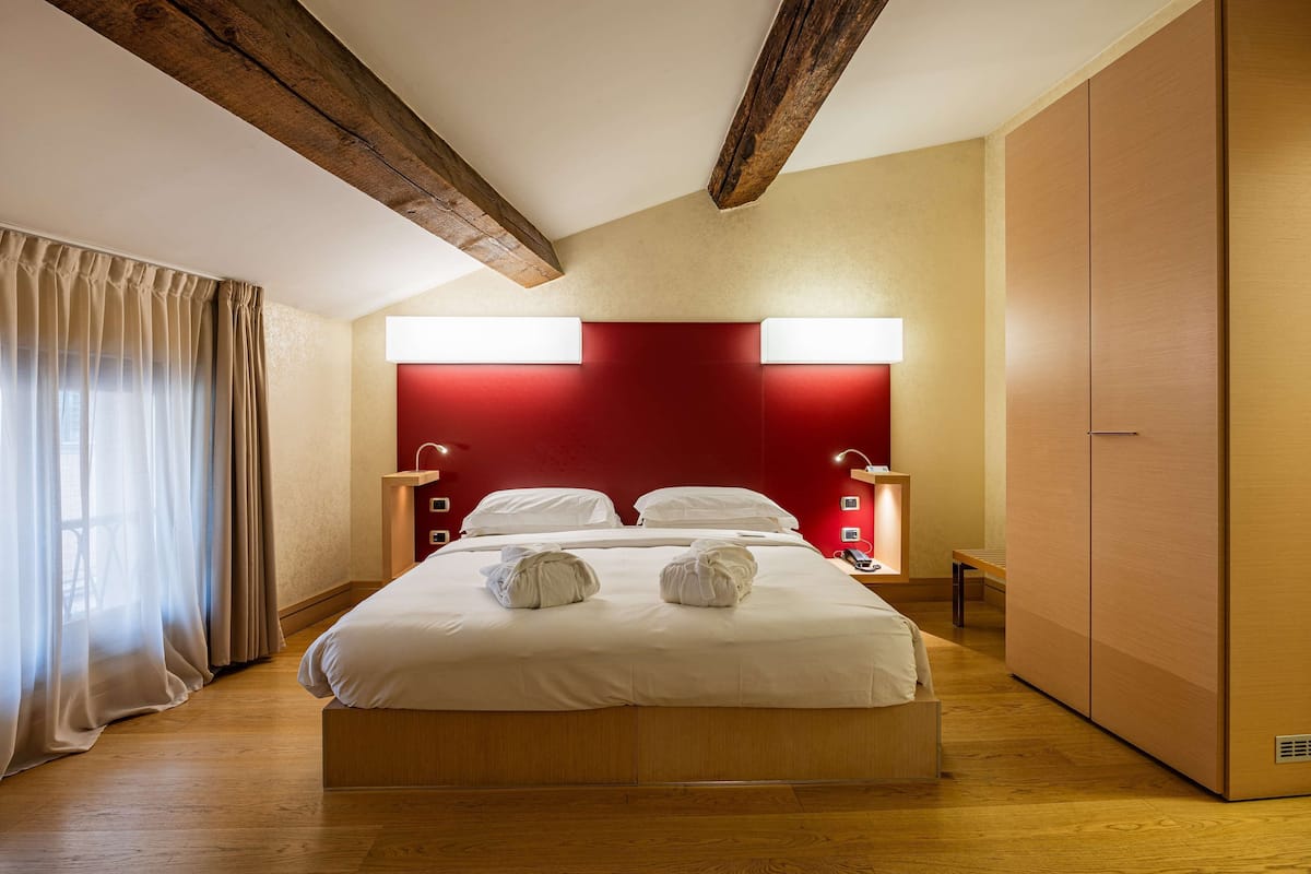 Quarto de hotel com teto de madeira, parede em destaque vermelha, cama grande com roupa de cama branca, duas luminárias de cabeceira e um guarda-roupa.