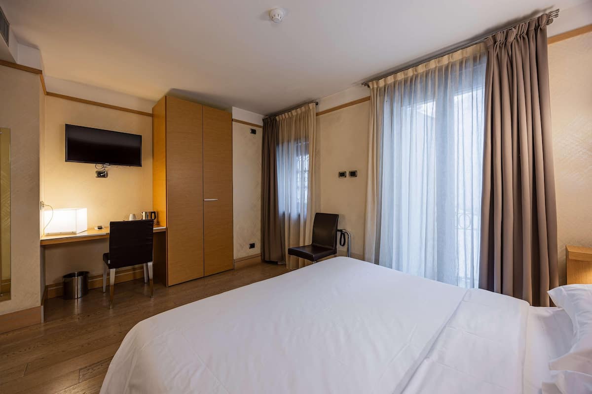 Quarto de hotel com uma cama grande, uma escrivaninha com cadeira, televisão na parede e cortinas nas janelas.