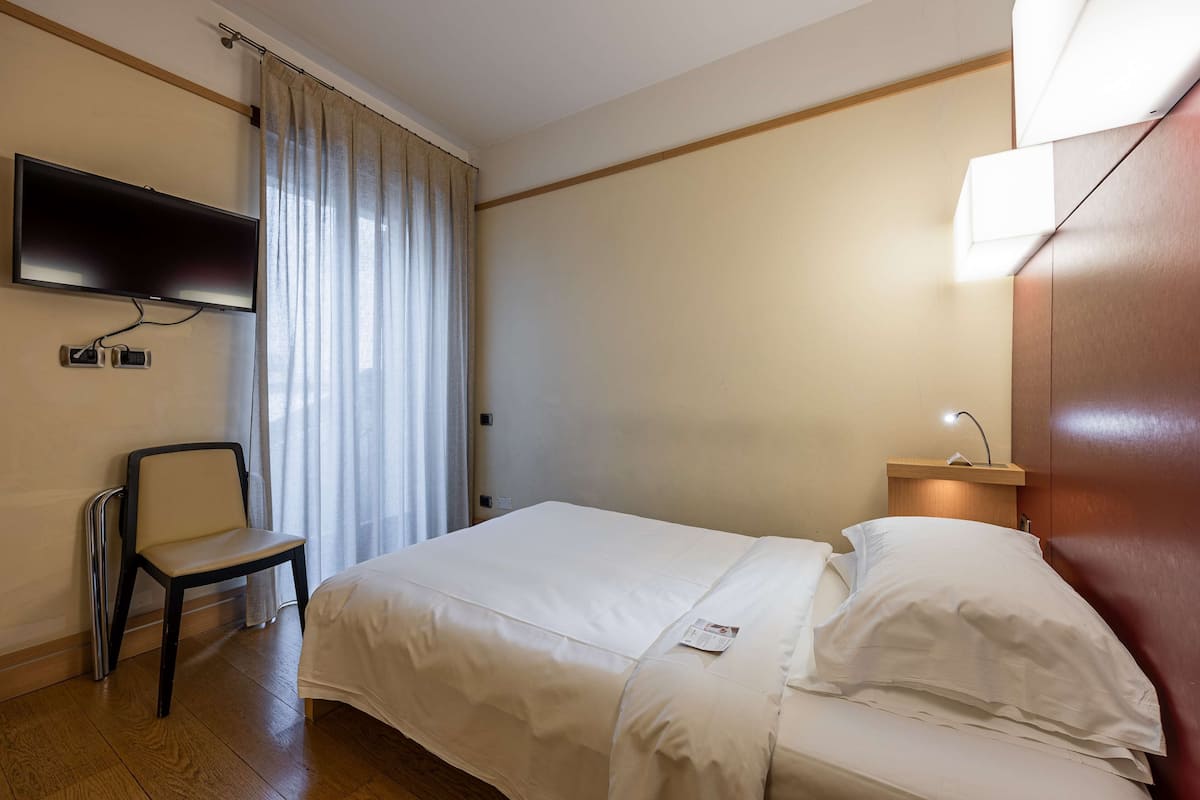 Quarto de hotel com uma cama, uma cadeira, uma televisão fixada na parede e uma mesa de cabeceira com abajur.