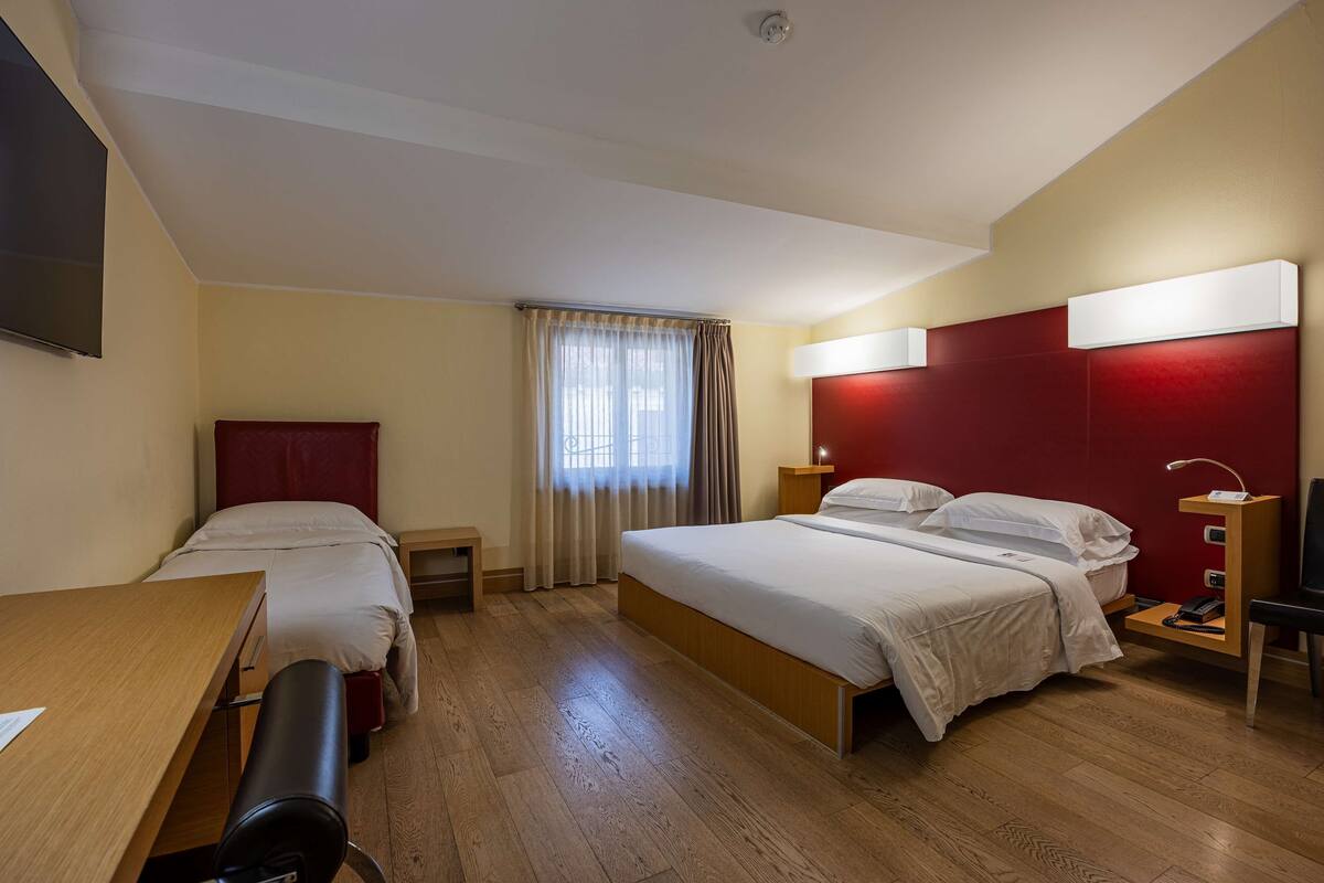 Quarto de hotel com duas camas, uma escrivaninha, uma cadeira, uma televisão e uma janela com cortinas.