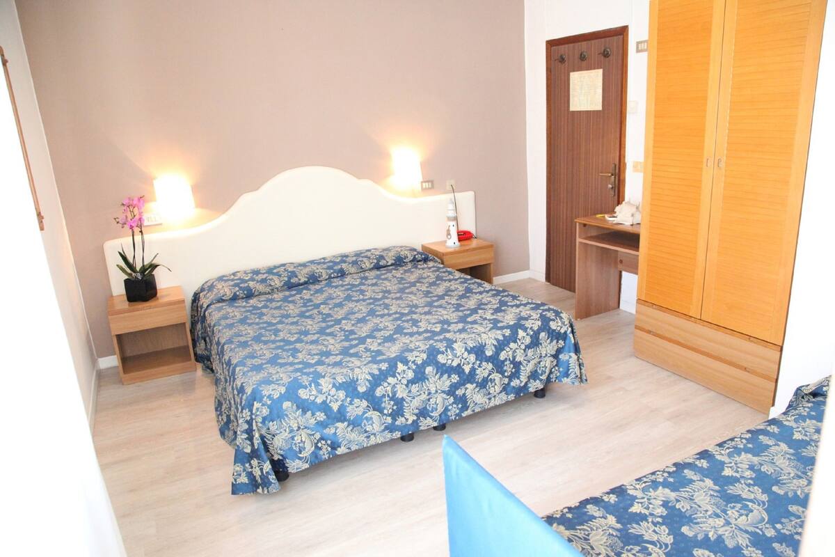 Quarto com cama, mesas de cabeceira, guarda-roupa e mesa de apoio com abajur.
