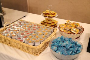 Mesa de buffet com diversos alimentos, incluindo potes de compotas, uma tigela de lanches embalados em azul e uma mesa escalonada com sobremesas.