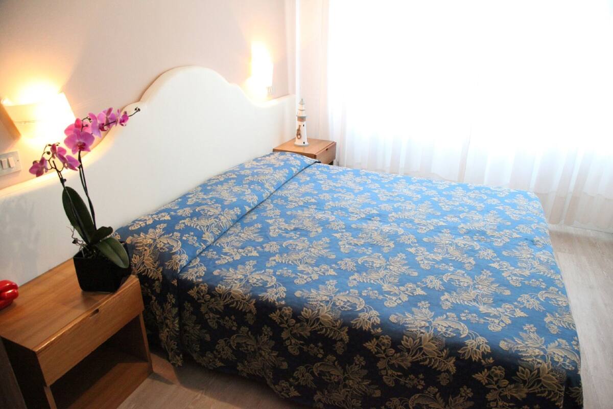 Quarto com cama, mesa de cabeceira, abajur e uma orquídea em vaso.
