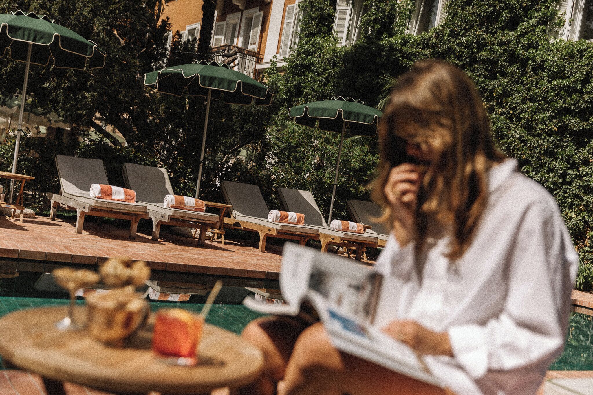 Área ao redor da piscina com espreguiçadeiras e guarda-sóis, uma mesa pequena com uma bebida, e uma pessoa lendo uma revista.