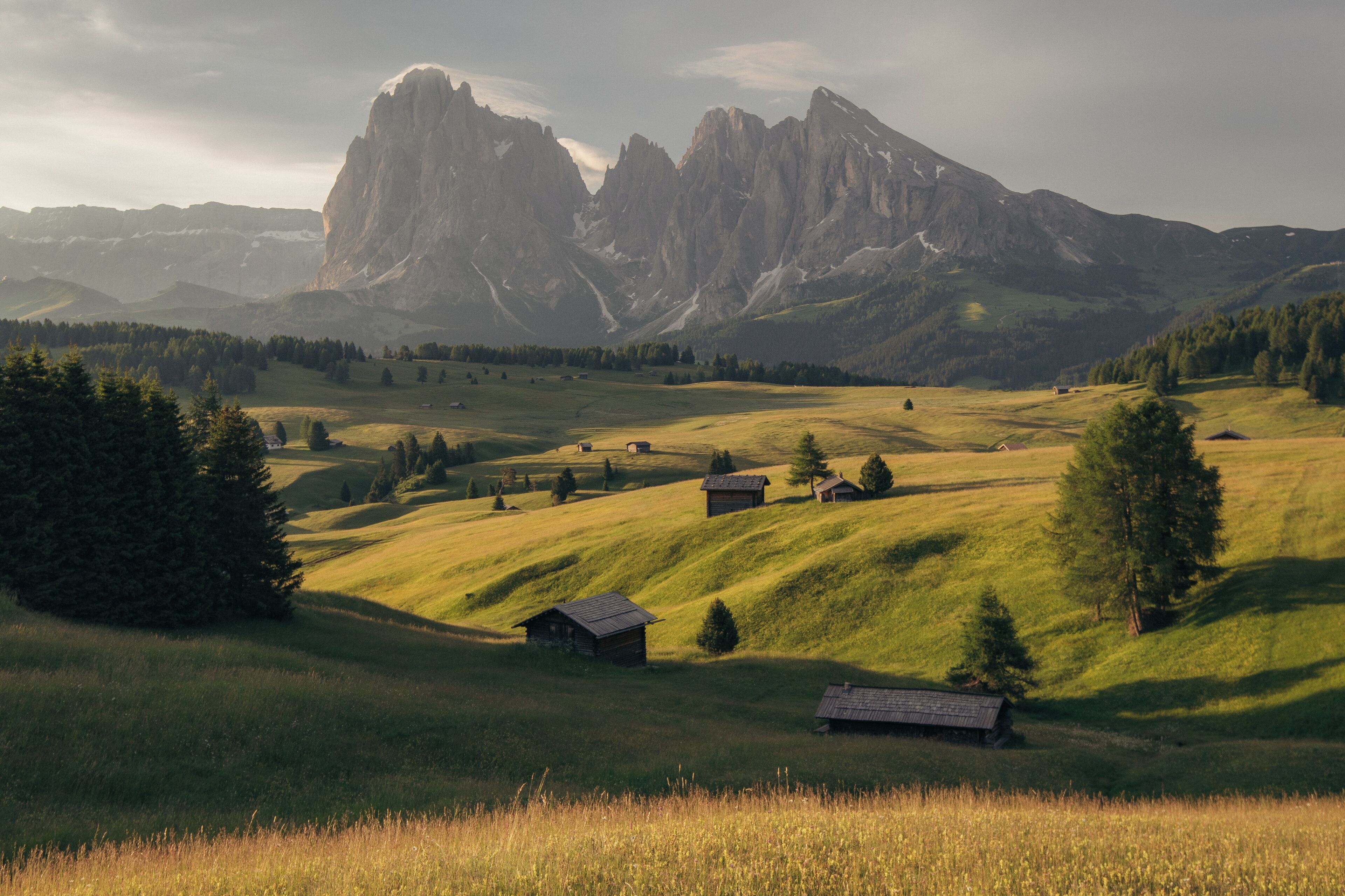 Paisagem montanhosa com colinas verdes ondulantes, árvores dispersas e alguns edifícios pequenos.