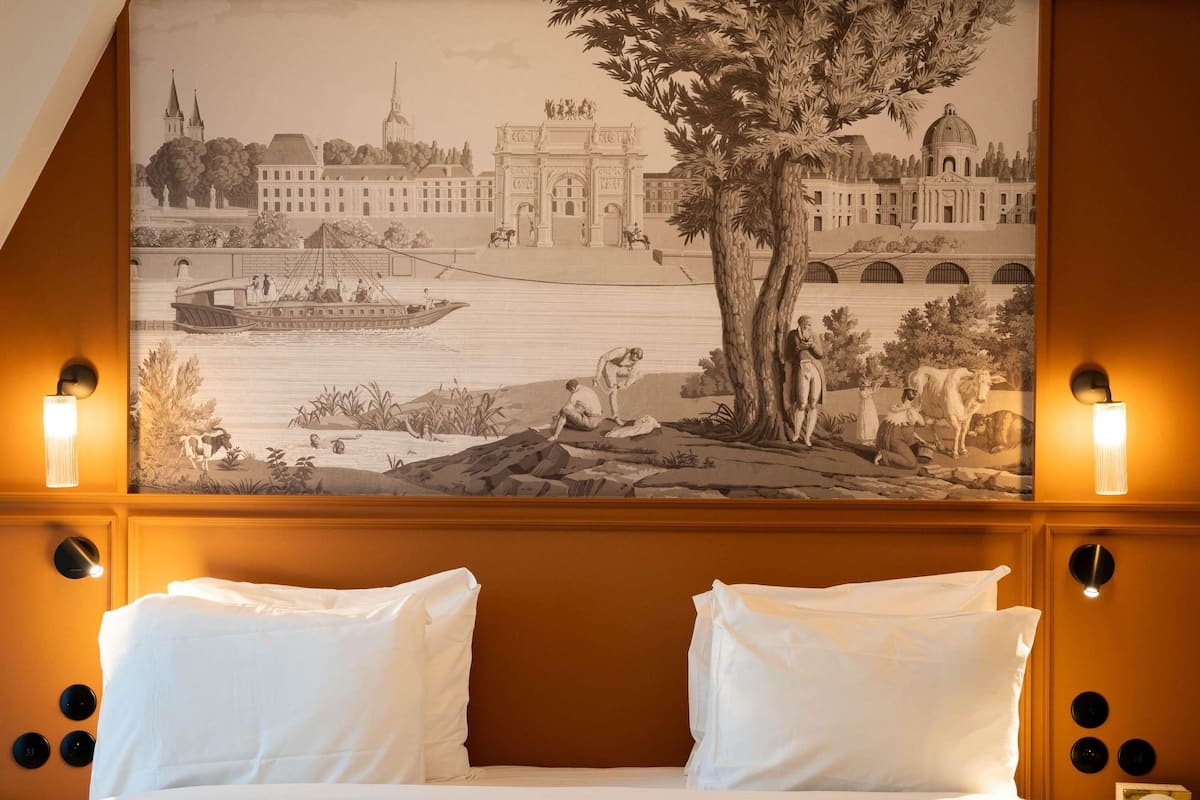 Quarto de hotel com um grande mural de uma paisagem urbana histórica, uma cama com lençóis brancos e arandelas na parede.
