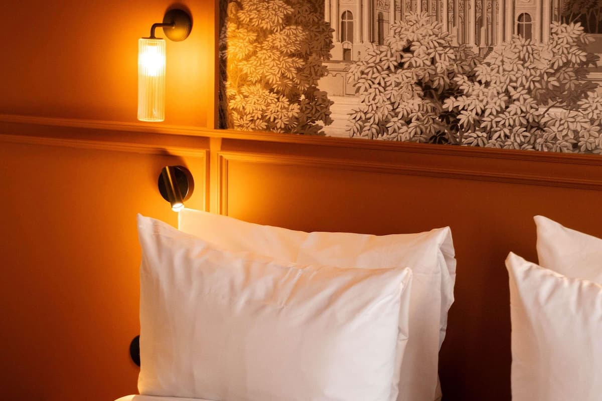Quarto de hotel com uma cama, almofadas brancas, luminária fixada na parede e painel decorativo.