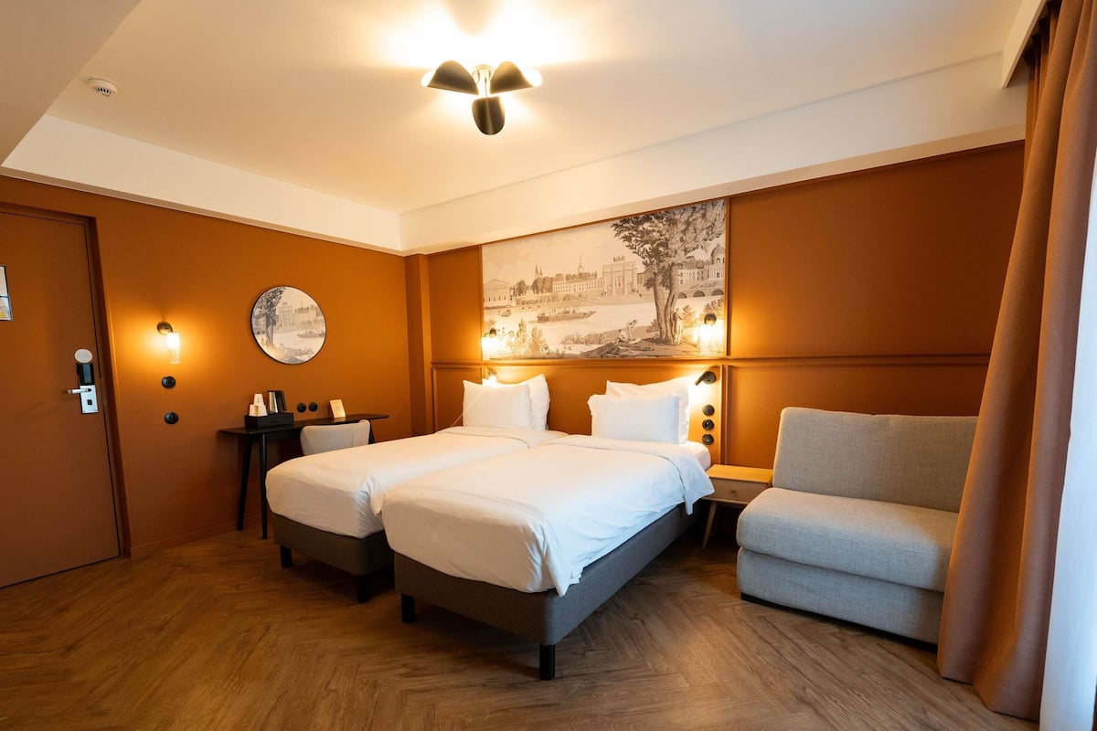 Quarto de hotel com duas camas, uma escrivaninha, uma cadeira e uma obra de arte na parede.