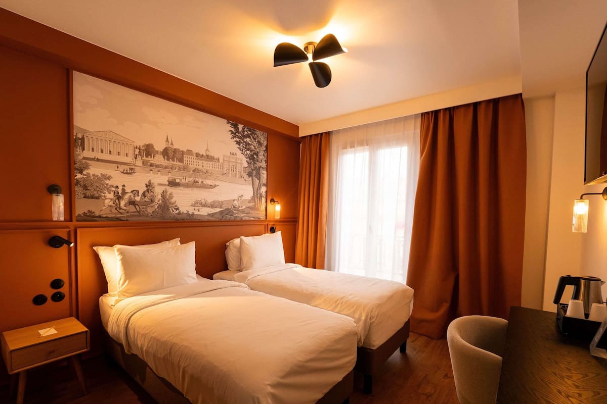 Quarto de hotel com duas camas, piso de madeira, um quadro na parede, uma mesa com cadeira e uma janela com cortinas.
