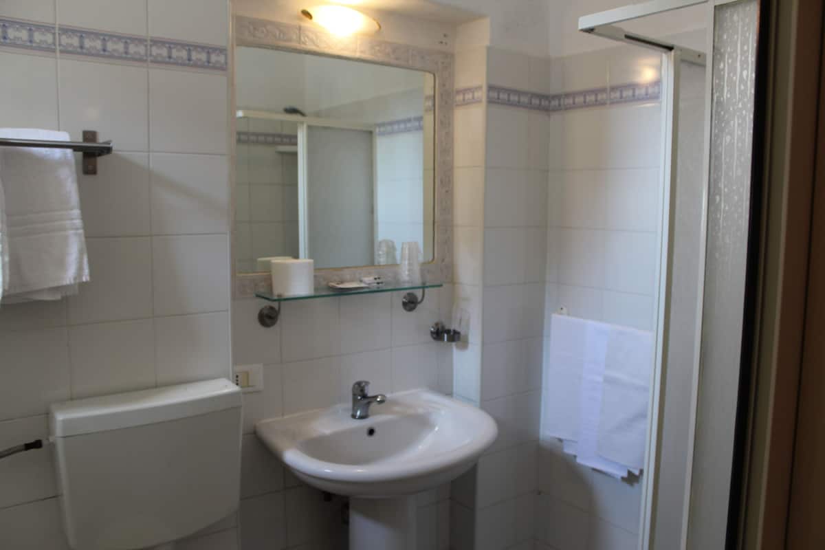 Casa de banho com lavatório branco, espelho, prateleira de vidro com produtos de higiene e barra de toalhas.