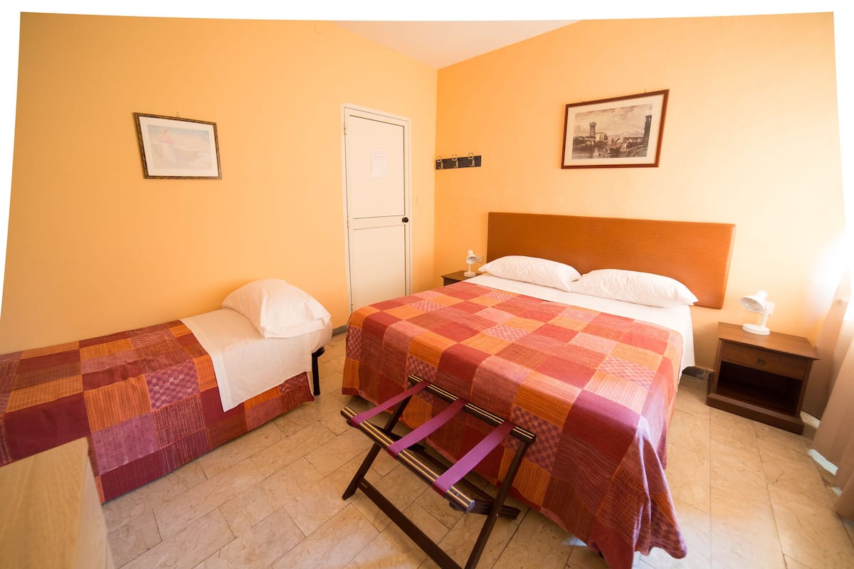 Quarto com duas camas, cabeceira de madeira, mesa de cabeceira com abajur e duas imagens emolduradas nas paredes.