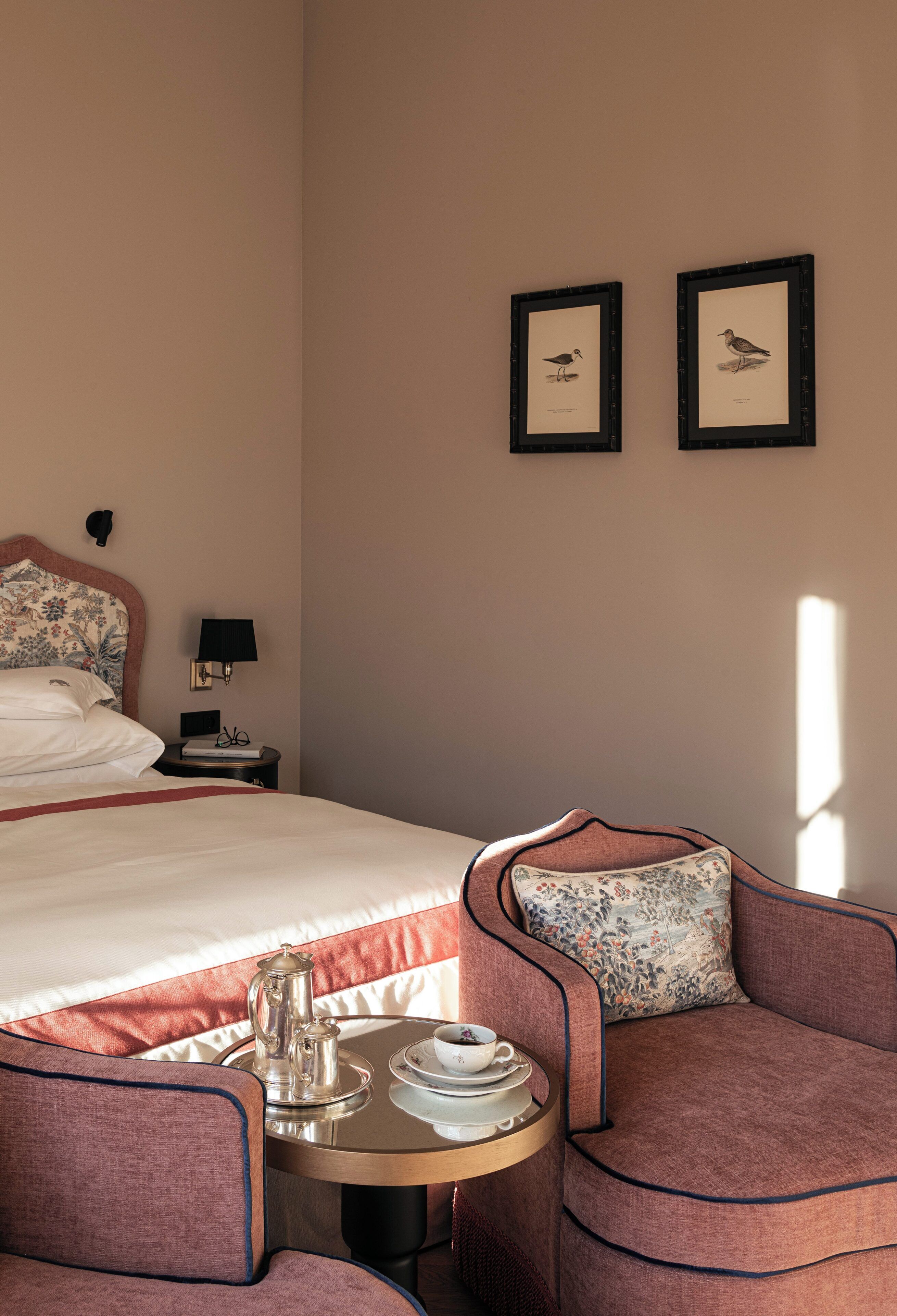 Quarto de hotel com cama, duas imagens de pássaros emolduradas, mesa de cabeceira com abajur, pequena mesa com bule e xícaras, e duas poltronas estofadas.