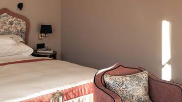 Quarto de hotel com cama, duas imagens de pássaros emolduradas, mesa de cabeceira com abajur, pequena mesa com bule e xícaras, e duas poltronas estofadas.