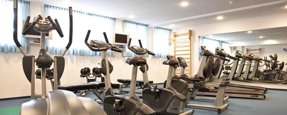 Uma academia com várias máquinas de cardio, uma televisão e um espelho grande.