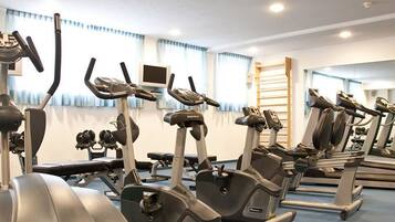 Uma academia com várias máquinas de cardio, uma televisão e um espelho grande.