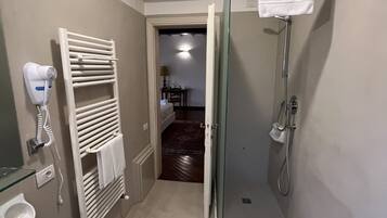 Casa de banho com cabine de chuveiro em vidro, toalha branca pendurada na porta do chuveiro, secador de cabelo na parede e uma pequena pia com espelho.