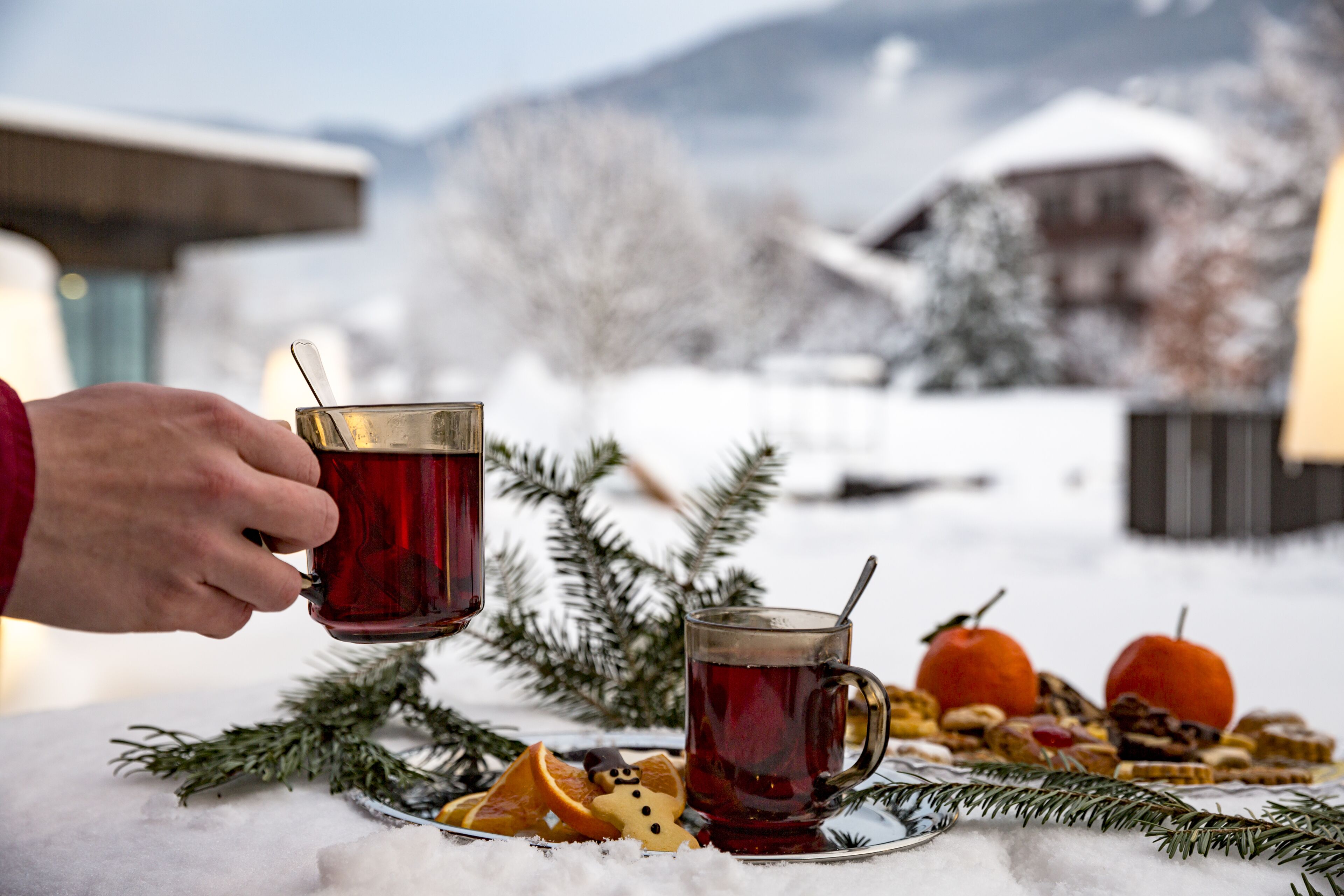 Duas chávenas de bebida vermelha sobre uma mesa coberta de neve, com uma decoração festiva incluindo bolachas e laranjas.
