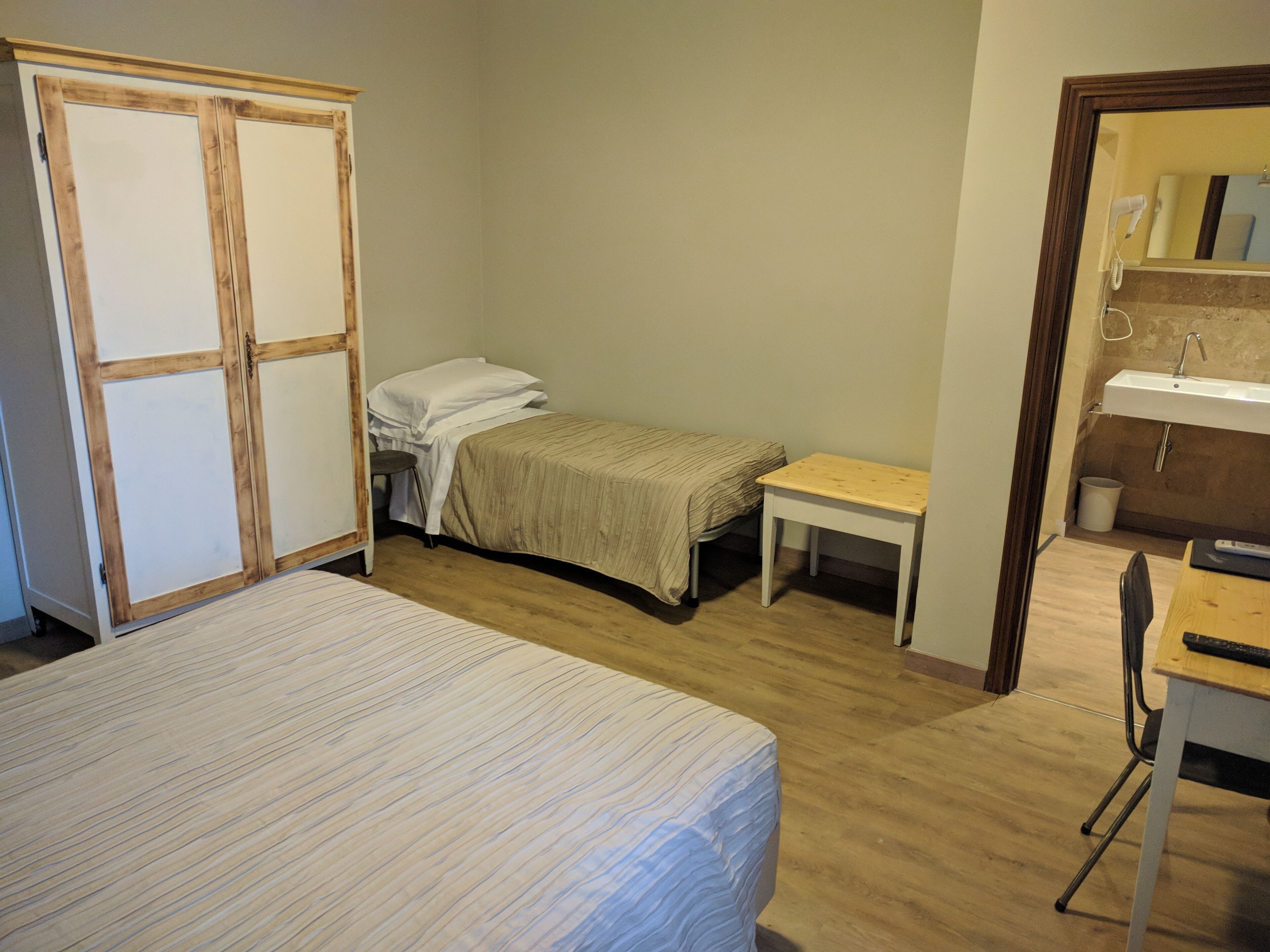 Quarto com cama, mesa de cabeceira pequena, cadeira, guarda-roupa e banheiro com pia e espelho.